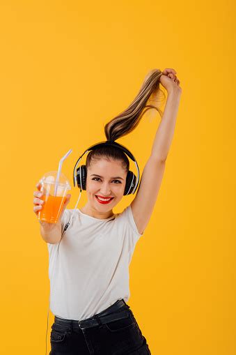 미소 아름다운 소녀는 카메라를 보고 머리 헤드폰입니다 흰색 셔츠를 입고 주스와 플라스틱 컵을 들고 노란색 배경에 고립 된 긍정적 인 얼굴 감정 복사 공간 귀여운에 대한 스톡
