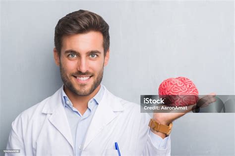 서 치아를 보여주는 자신감 미소로 웃 고 행복 한 얼굴로 뇌를 들고 회색 그런 지 벽에 잘생긴 젊은 의사 심리학자 남자 Mental Wellbeing에 대한 스톡 사진 및