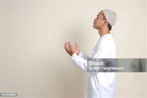 해골 모자를 쓴 흰 셔츠를 입은 매력적인 아시아 무슬림 남자의 초상화가 손을 들고 간절히 기도하고 있다 회색 배경에 격리된