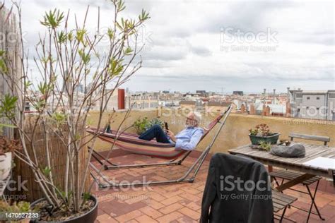 회색 머리와 턱수염을 기른 성숙한 사업가가 파란 셔츠와 청바지를 입고 테라스에서 스마트폰을 들고 해먹에 누워 있다 60 69세에 대한 스톡 사진 및 기타 이미지 Istock