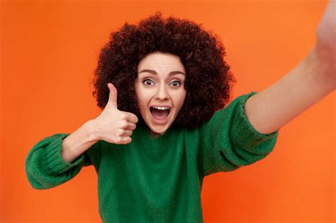 녹색 캐주얼 스타일의 스웨터를 입고 셀카를 찍고 행복을 표현하며 카메라에 엄지 손가락을 드러내는 아프로 헤어 스타일을 입은 흥분한 여성 엄지손가락을 위로에 대한 스톡 사진