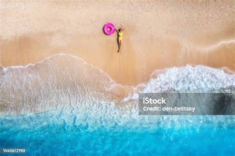 해질녘에 파도가 있는 바다 근처의 모래 해변에 분홍색 수영 반지를 끼고 누워 있는 아름다운 젊은 여성의 공중 전망 그리스 레프카다 섬에서의 여름 휴가 날씬한 소녀 맑은 푸른