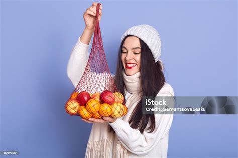 만다린과 사과를 보고 손에 그물 핸드백을 들고 좋은 분위기에 있고 과일을 좋아하고 혼자 시간을 보내고 빨간 입술을 갖는 쾌활한 외모의 갈색 머리의 초상화 새해 분위기 감귤에