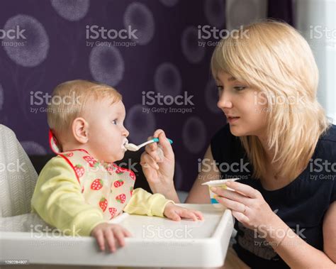 엄마는 항아리에서 아기 흰 요구르트를 공급합니다 아이는 높은 의자에 앉아있다 내 얼굴은 요구르트로 얼룩져 있습니다 앞치마가