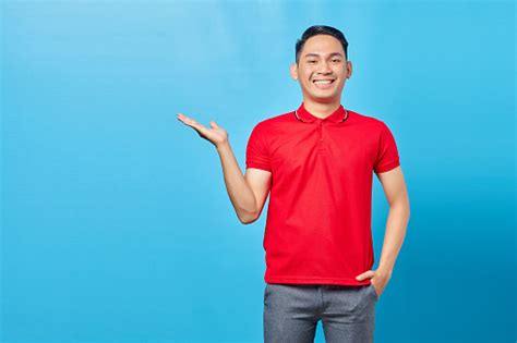 빨간 셔츠를 입은 쾌활한 아시아 청년이 파란 배경에 고립 된 손바닥으로 복사 공간을 가리키고 서있는 초상화 25 29세에 대한 스톡 사진 및 기타 이미지 Istock
