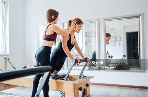 여성 필라테스 강사는 스포츠 운동 체육관 홀에서 개혁기를 사용하여 Abs 근육 근력 운동을 하는 자세 조정을 돕습니다 활동적인 사람들 훈련 요가 수업 개념 2명에 대한 스톡