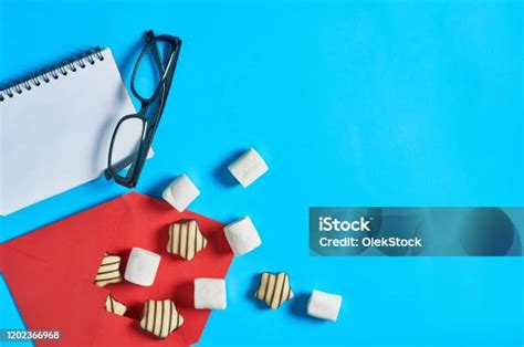 흩어져있는 과자 사탕 광학 안경 근처의 마시멜로 빈 메모장이있는 빨간 종이 봉투가 파란색 책상 위에 놓여 있습니다 사랑 편지 개념입니다 텍스트의 공백입니다 맨 위 보기