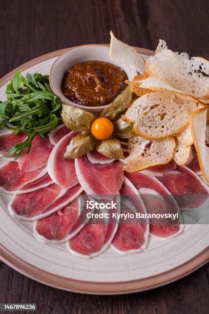 밀 바게트 빵 칩 루꼴라 매실 소스를 곁들인 송아지 카르파치오 음식은 가볍고 둥근 세라믹 접시에 있습니다 접시는 갈색 나무