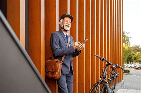 휴대전화를 들고 미소 짓는 노인 통근자가 벽가에 서서 자전거 근처에 서서 생각에 잠긴 채 올려다보고 있습니다 60 69세에 대한 스톡 사진 및 기타 이미지 Istock