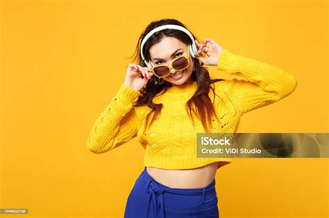 스웨터에 웃는 여자 파란 바지 선글라스 노란색 오렌지 배경에 고립 된 카메라를 찾고 헤드폰 듣기 음악 사람들은 진심으로 감정 라이프 스타일 개념입니다 복사 공간을 모의