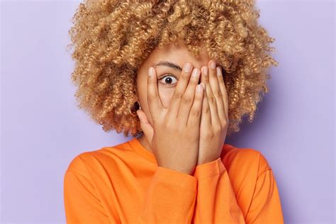곱슬곱슬한 금발 머리를 한 겁먹은 여성이 손바닥으로 얼굴을 가리고 손가락을 통해 얼굴을 가립니다 보라색 배경 위에 격리된 주황색 점퍼를 입고 무서운 것을 숨기려고 합니다
