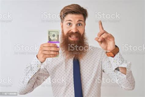 젊은 빨간 머리 아일랜드어 사업가 는 행복한 얼굴 번호 하나 와 손가락을 가리키는 아이디어 또는 질문으로 놀란 고립 된 흰색 배경 위에 서 있는 달러를 들고 가리키기에 대한