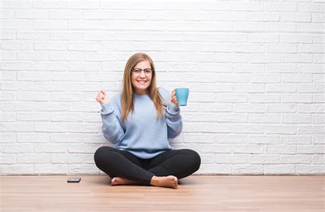 젊은 성인 여자 커피 자랑 비명과 승리와 성공을 매우 흥분된 하 고 축 하의 흰색 벽돌 벽 Driking 컵 바닥에 앉아 감정을 응원 감정에 대한 스톡 사진 및 기타 이미지