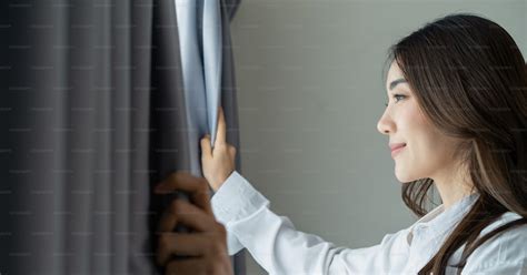 아시아의 아름다운 여자가 아침에 창문에 접근하고 커튼을 엽니다 흰 잠옷을 입은 젊은 매력적인 소녀는 집에서 침실에서 일찍 일어난 후 밖을 내다보고 일출을 보는 것이