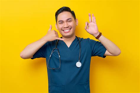 파란색 유니폼을 입고 청진기를 착용한 행복한 전문 아시아 남성 의사 또는 간호사가 노란색 배경에 격리된 나에게 다시 전화하고 괜찮다는 신호를 보내는 것과 같은 제스처를