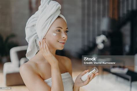 머리에 흰색 목욕 수건을 가진 아름다운 젊은 유럽 여성은 얼굴 크림과 바디 로션을 적용하고 샤워 후 피부 관리 절차를 즐기고