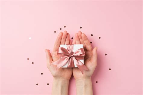 1인칭 탑 뷰 사진 세인트 발렌타인 데이 장식 젊은 여성의 손은 손바닥에 분홍색 리본 리본이 달린 작은 흰색 선물 상자를 들고 있으며 고립 된 파스텔 핑크 배경에 빛나는