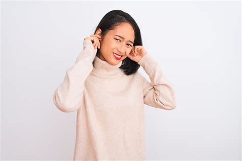 시끄러운 음악의 소음에 대한 성가신 표정으로 손가락으로 귀를 덮는 고립 된 흰색 배경 위에 서있는 터틀넥 스웨터를 입은 젊은 중국 여성 청각 장애인 개념입니다 가리기에 대한