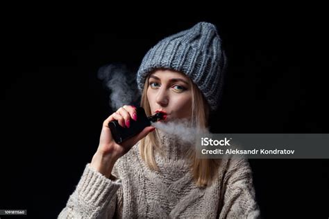 여 자가 니트 스웨터와 모자에 금발 전자 담배 Vape 매니큐어와 그녀의 손에 보유 하 고 그녀의 입술에 빨간 립스틱으로 그린 연기 근 데 재미에 대한 스톡 사진 및 기타