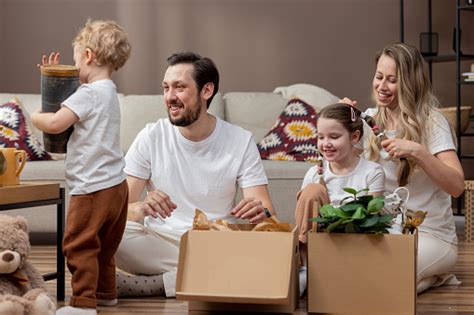 두 명의 어린 자녀를 둔 부모는 새 아파트로 이사하고 골판지 상자를 풀고 물건을 꺼내고 달콤한 아들은 꽃병을 나르고 엄마는 딸의 끈을 꼰다 30 34세에 대한 스톡 사진 및