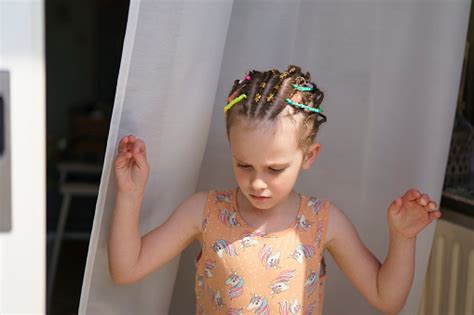 형형색색의 작은 땋은 머리를 한 작고 귀엽고 슬픈 소녀가 방 입구의 하얀 커튼을 배경으로 서 있습니다 어린아이와 같은 슬픔과