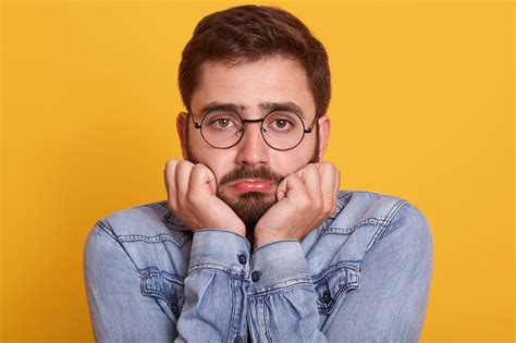 불행한 기분을 상하게 한 젊은 수염 남성 곡선은 입술을 낮추고 턱 아래에 손을 유지하며 노란색 스튜디오 배경을 배경으로 포즈를 취합니다 세련된 데님 재킷과 안경을 입고 백인