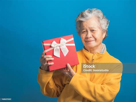 빨간 선물 상자를 들고 파란 배경에 서서 미소를 지으며 카메라를 바라보는 쾌활한 선배 여성 선물에 대한 스톡 사진 및 기타 이미지 Istock
