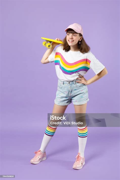 스튜디오에서 보라색 파스텔 벽 배경에 고립 된 옆을 찾고 노란 스케이트 보드를 들고 서 있는 생생한 옷에 웃는 십 대 소녀 사람들은 진심으로 감정 라이프 스타일 개념입니다