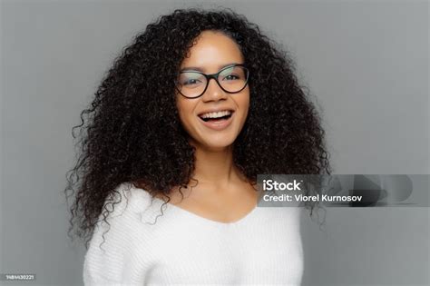 낙관적인 곱슬머리 여성의 솔직한 샷은 긍정적으로 웃고 기분이 좋고 건강한 피부를 가지고 있고 투명한 안경을 통해 보고 흰색 스웨터를 입고 회색 배경 위에 고립되어 있습니다