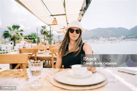 선글라스와 모자를 쓴 젊은 여성 여성이 바다와 산을 배경으로 한 거리의 야외 해변 카페 레스토랑 커피숍에서 커피를 마신다