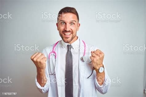 고립 된 배경위에 분홍색 청진기를 입은 젊은 잘 생긴 소아과 의사 남자는 팔을 들고 눈을 뜨고 성공을 축하하고 놀랐습니다 우승자 컨셉 건강관리와 의술에 대한 스톡 사진 및