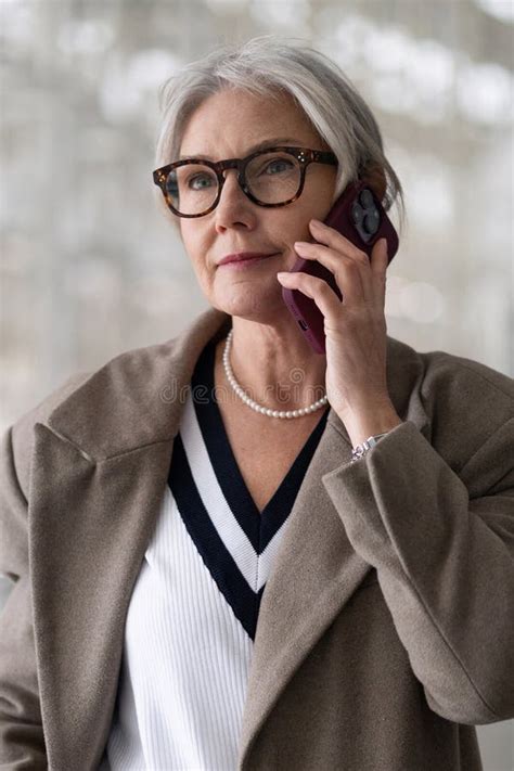 An Older Woman In Glasses Talks On Her Phone While Dressed In A Stylish Coat And Pearl Necklace