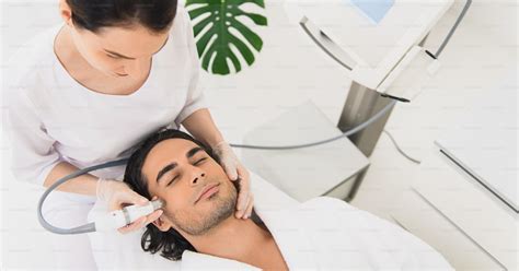 숙련 된 미용사는 캐비테이션 장비로 남성 얼굴을 정화하고 있습니다 남자는 거짓말을하고 긴장을 풀고 있습니다 그의 눈은 기쁨으로 감겨있다 사진 Unsplash의 뷰티