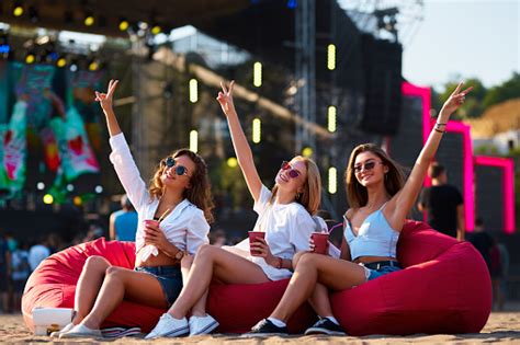 해질녘 팥 주머니를 타고 음료를 마시며 환호하고 모래 해변 축제에서 라이브 음악을 즐기는 즐거운 소녀들 캐주얼한 여름 복장 선글라스 활기찬 축제 분위기 바다 배경 Live