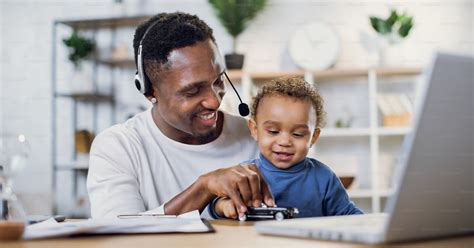 헤드셋을 쓴 아프리카계 미국인 아버지는 귀여운 아들과 놀고 현대 노트북에서 온라인 회의를 하고 있다 젊은 남자는 아기와 함께 집에 앉아 거리를 두고 일하고 있다 사진