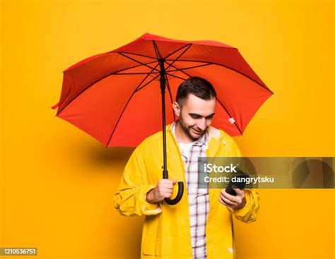 빨간 우산 사용 휴대 전화 오렌지 배경을 통해 고립 된 노란색 비옷에 젊은 잘 생긴 수염 남자 전화에 대한 스톡 사진 및 기타 이미지 Istock