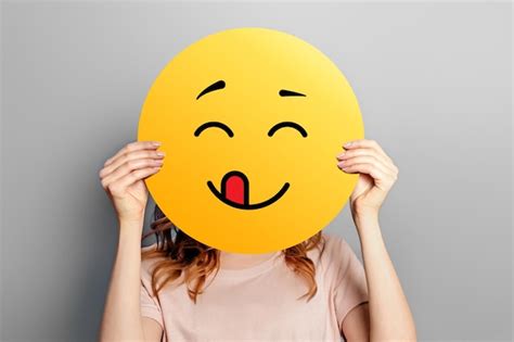 Emoticono Lamiendo La Chica Sostiene Una Carita Amarilla Con La Lengua Aislada En Un Fondo Gris