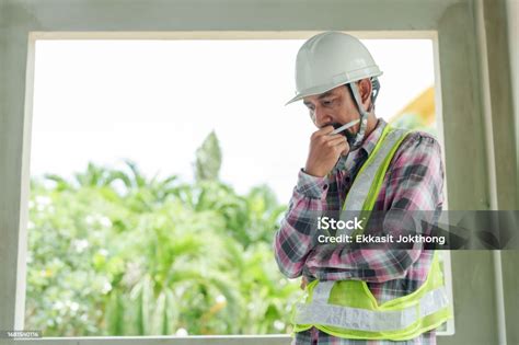 턱수염을 기른 아시아계 건설 노동자가 서서 작업 계획에 대해 생각하고 있다 녹색 반사 조끼와 안전 헬멧을 착용한 남자는 건설 현장에서 일하는 동안 혼란스러운 얼굴을 하고