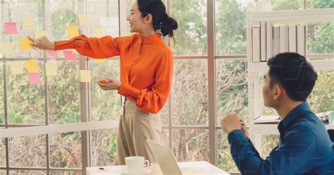 비즈니스 사람들은 사무실의 프로젝트 계획 보드에서 일하고 동료 친구와 대화하여 프로젝트 개발을 분석합니다 그들은 유리 벽에 게시 된 스티커 메모를 사용하여 정리합니다