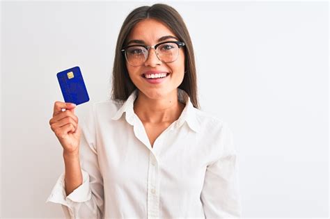 고립된 흰색 배경 위에 신용 카드를 들고 안경을 쓰고 행복한 얼굴을 하고 치아를 보여주는 자신감 있는 미소로 미소 짓는 젊은 사업가 건강한 생활방식에 대한 스톡 사진 및