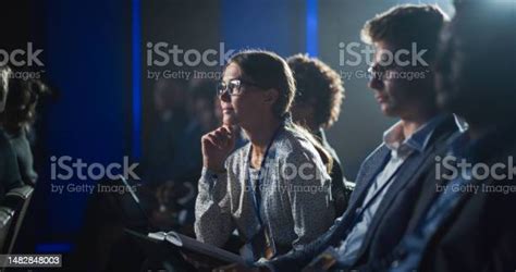 비즈니스 컨퍼런스에서 붐비는 청중에 앉아있는 젊은 여성 여성 참석자가 중요한 글머리 기호를 노트북에 적어 둡니다 성공적인 사업가들과 함께 강당에서 기조 연설 기자 회견에