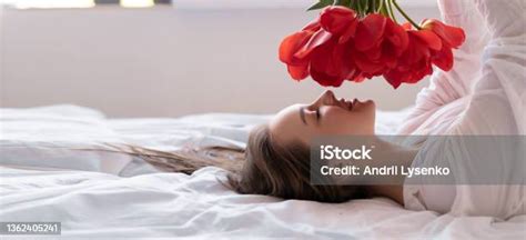 젊은 아름다운 백인 여성이 하얀 셔츠를 입고 침대에 누워 빨간 튤립을 손에 들고 향기를 흡입합니다 20 24세에 대한 스톡 사진 및 기타 이미지 Istock