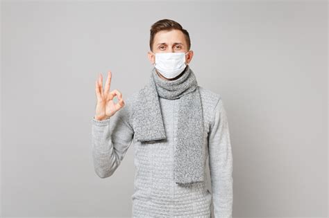 회색 스웨터에 젊은 남자 스튜디오에서 회색 배경에 고립 된 확인 제스처를 보여주는 멸 균 얼굴 마스크 스카프 건강 한 라이프 스타일 아픈 질병 치료 추운 계절 개념입니다