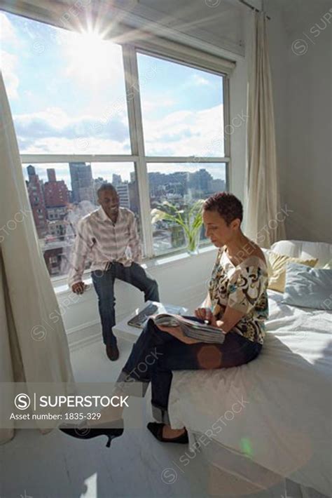 Side Profile Of A Woman Sitting On A Bed And A Man Sitting At The Window Sill SuperStock