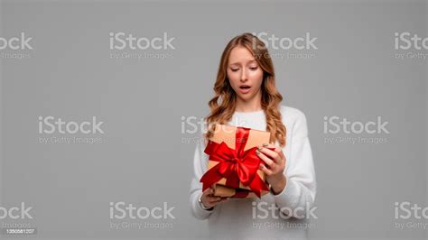 충격적인 표정으로 선물 상자 안을 들여다보는 흰색 스웨터를 입은 젊은 여성이 비밀리에 선물을 풀고 회색 배경 위에 서서 텍스트를 위한 복사 공간을 가지고 서 있습니다 25