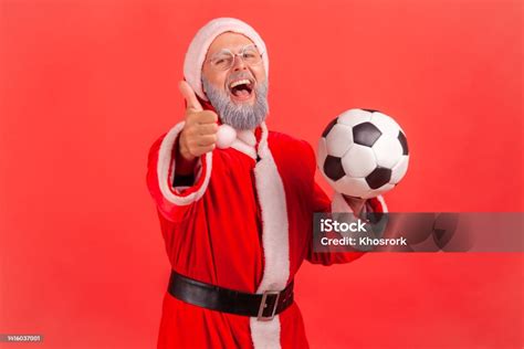 회색 수염을 가진 웃는 노인이 축구공이 달린 산타클로스 의상을 입고 엄지 손가락을 들고 행복한 표정으로 카메라를 바라보는 모습 축구에 대한 스톡 사진 및 기타 이미지