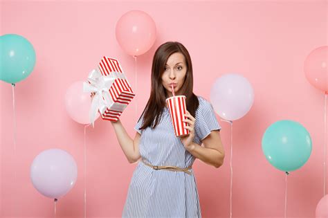 블루 드레스 선물 빨간색 상자를 들고 재미 있는 아름 다운 여자의 초상화 선물 마시는 소 다 또는 콜라 파스텔 핑크 배경에 플라스틱 컵에서 화려한 공기 풍선 생일 휴가 파티