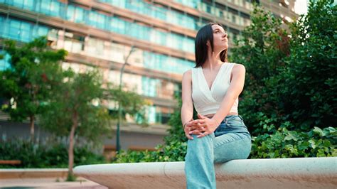 주근깨와 짙은 느슨한 머리카락을 가진 젊은 여성이 흰색 상단과 청바지를 입고 현대 도시의 전망을 즐기고 있습니다 긴 속눈썹을 가진 소녀는 현대 건물 배경의 공공 정원에 앉아