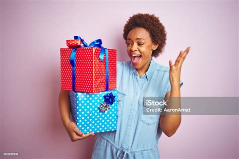 분홍색 고립 된 배경을 통해 생일 선물을 들고 아프리카 계 미국인 여성은 매우 행복하고 흥분 큰 미소와 손을 들고 비명을 지르는 승리를 축하하는 우승자 표정 공휴일에 대한