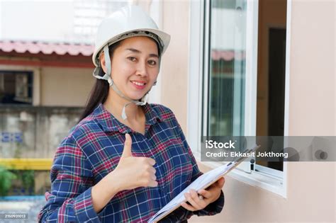 쾌활한 아시아 집 검사관이 엄지 손가락을 치켜 올리고 클립 보드를 들고 흰 헬멧을 쓰고 집 창문 옆에 서서 웃고 있습니다 검사관이 완공된 집을 검사합니다 30 34세에 대한